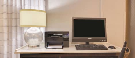 Um espaço de trabalho em hotel, com um computador de mesa, uma impressora, uma luminária e uma cadeira.