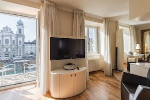Quarto de hotel moderno com televisão de tela plana, um armário redondo, área de refeições com mesa e cadeiras, e vista para um edifício histórico.