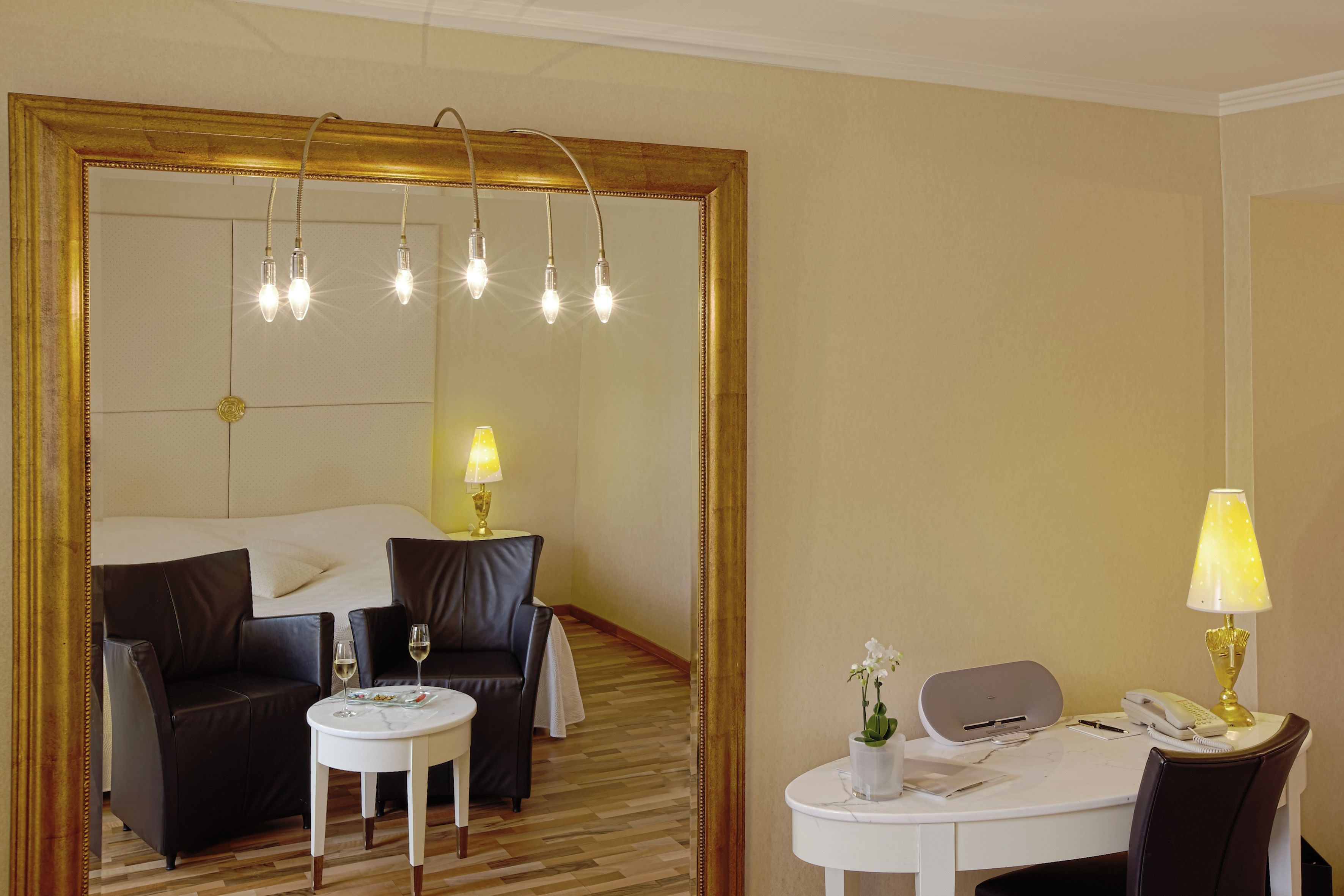 Quarto de hotel com cama, área de estar com mesa e cadeiras, escrivaninha com telefone e luminária, e espelho com moldura dourada.