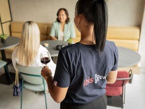 Uma pessoa vestindo uma camisa azul escura com a inscrição 'ibis Crew', segurando uma taça de vinho, sentada em uma mesa redonda com outra pessoa vestindo uma camisa verde clara.