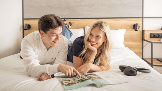 Um casal numa cama com uma revista, um bule de chá e uma mesa pequena com um abajur.