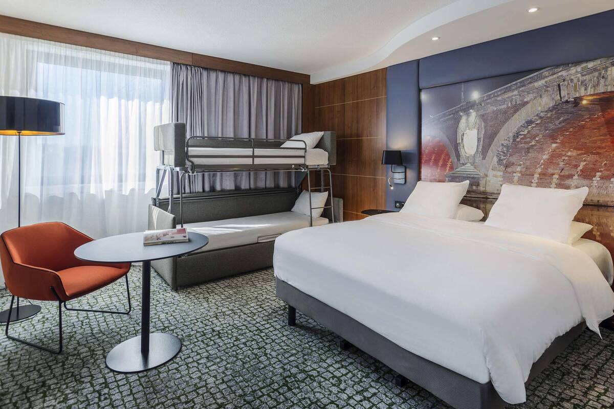 Quarto de hotel com uma cama beliche, uma cama de solteiro, uma mesa redonda, uma cadeira e um grande mural na parede.