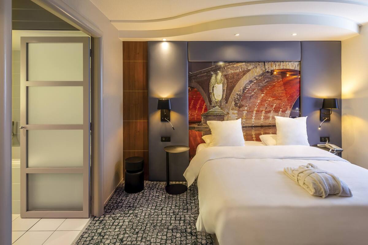 Quarto de hotel com uma cama, duas luminárias fixadas na parede, um quadro abstrato grande e uma porta de vidro que dá para um banheiro.