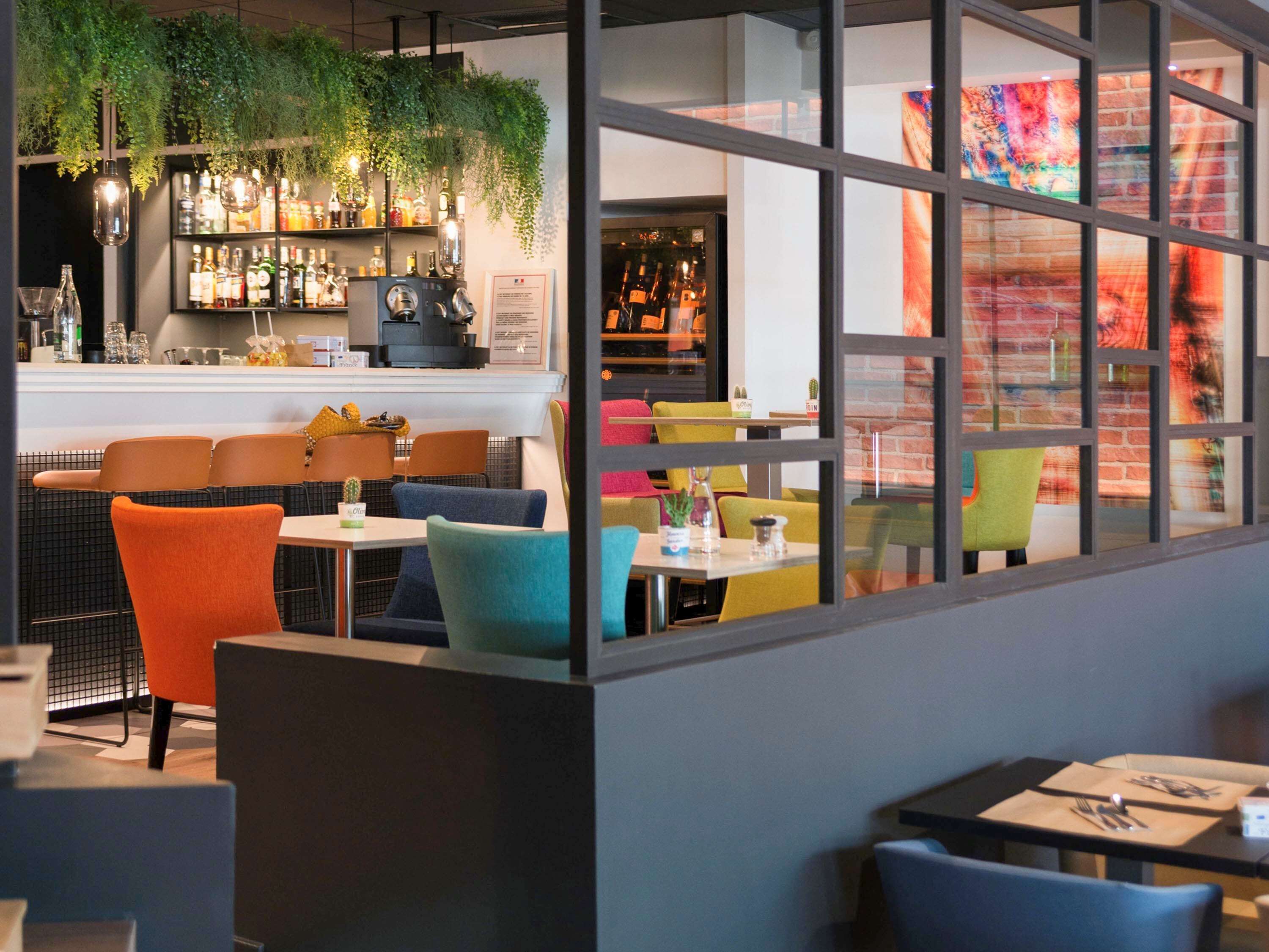 Uma área de bar com diversos tipos de garrafas e uma máquina de café, rodeada por assentos coloridos e uma planta pendurada no teto.