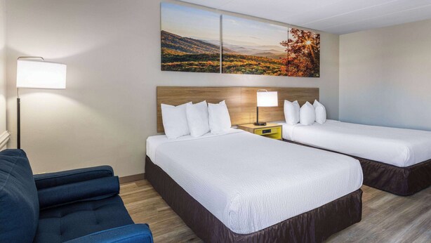 Quarto de hotel com duas camas, uma poltrona azul, uma mesinha de cabeceira com abajur e um quadro pendurado na parede.