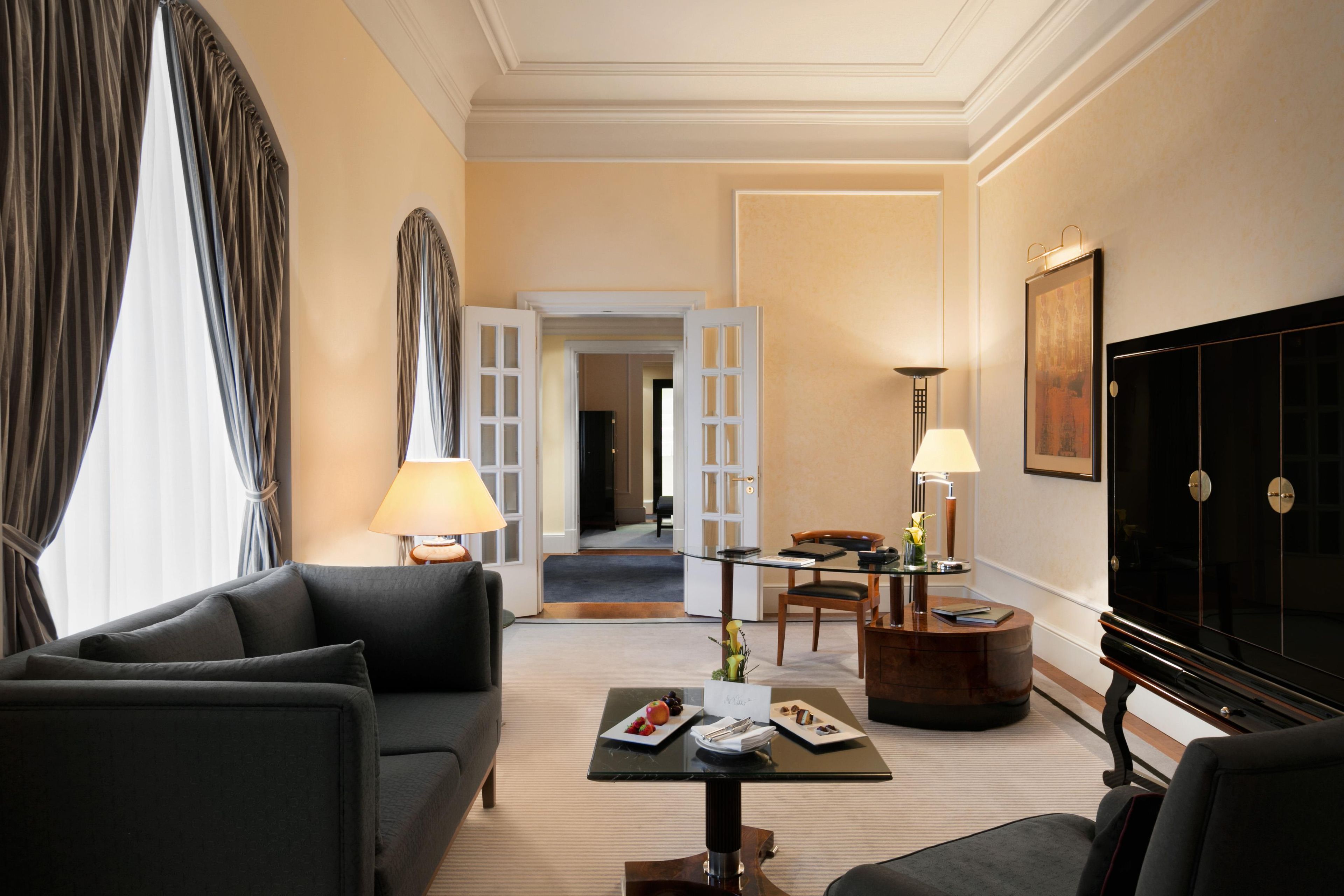 Quarto de hotel com sofá, mesa de centro com controle remoto, mesa de jantar com prato de comida e televisão na parede.