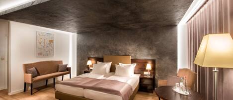Quarto de hotel moderno com uma cama grande, um sofá, uma mesinha de centro com uma luminária e um quadro na parede.