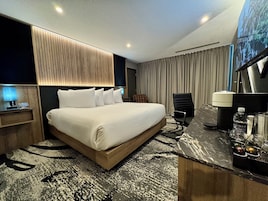 Quarto de hotel moderno com uma cama grande, uma mesa com cafeteira, uma cadeira e uma janela ampla com cortinas.