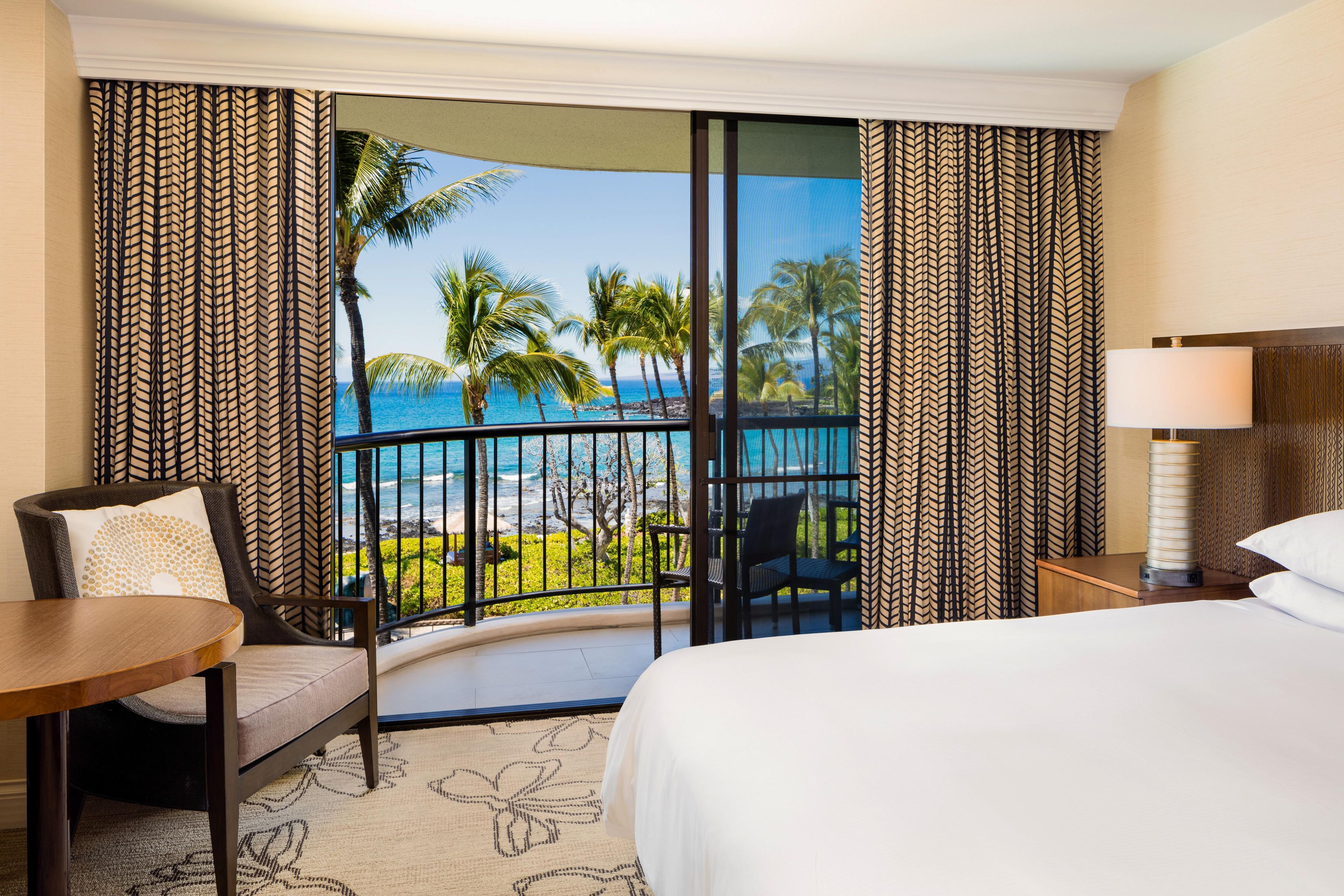 Quarto de hotel com cama, poltrona, mesinha e vista para o oceano com palmeiras.