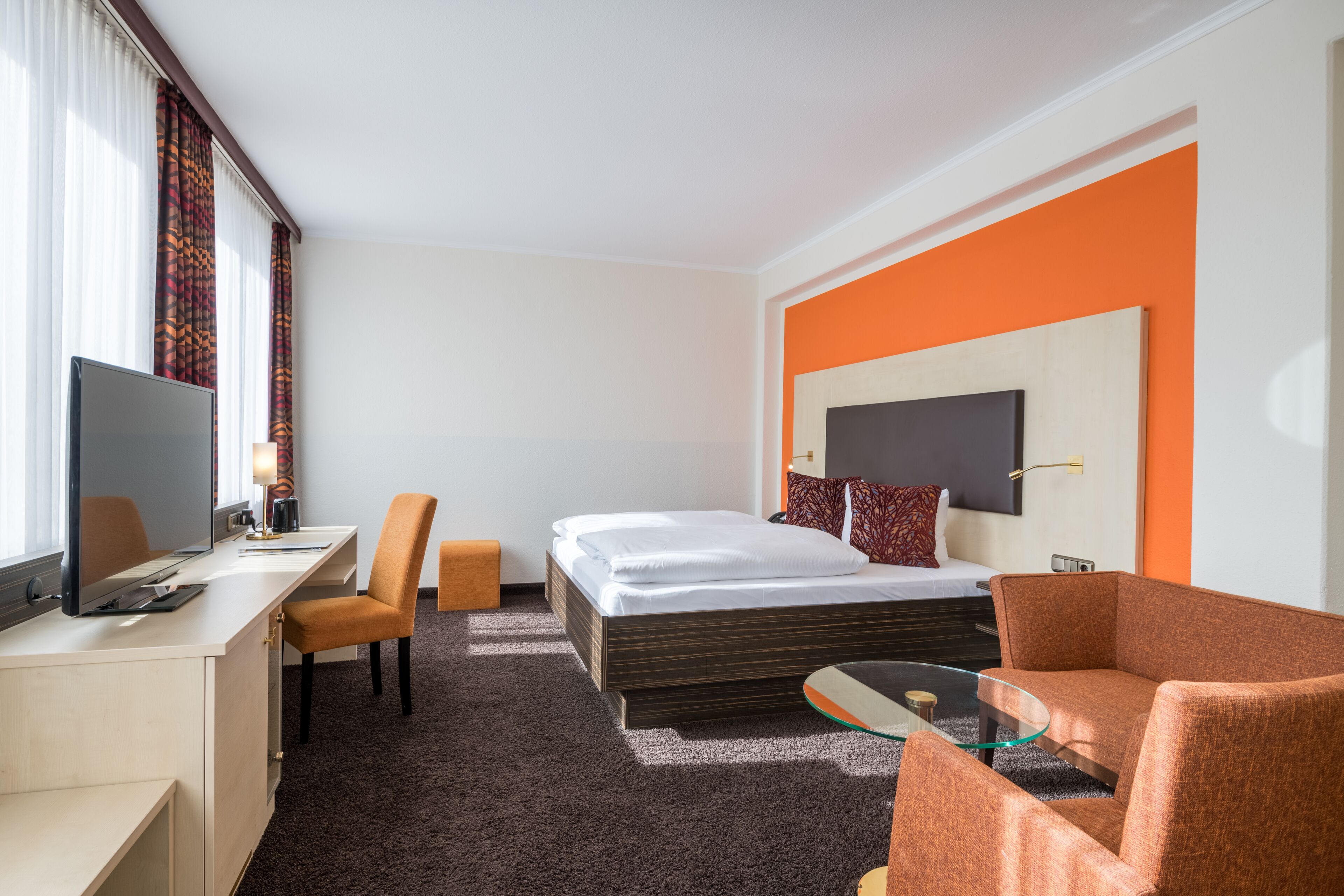 Quarto de hotel com cama, escrivaninha, poltrona, sofá e televisão.