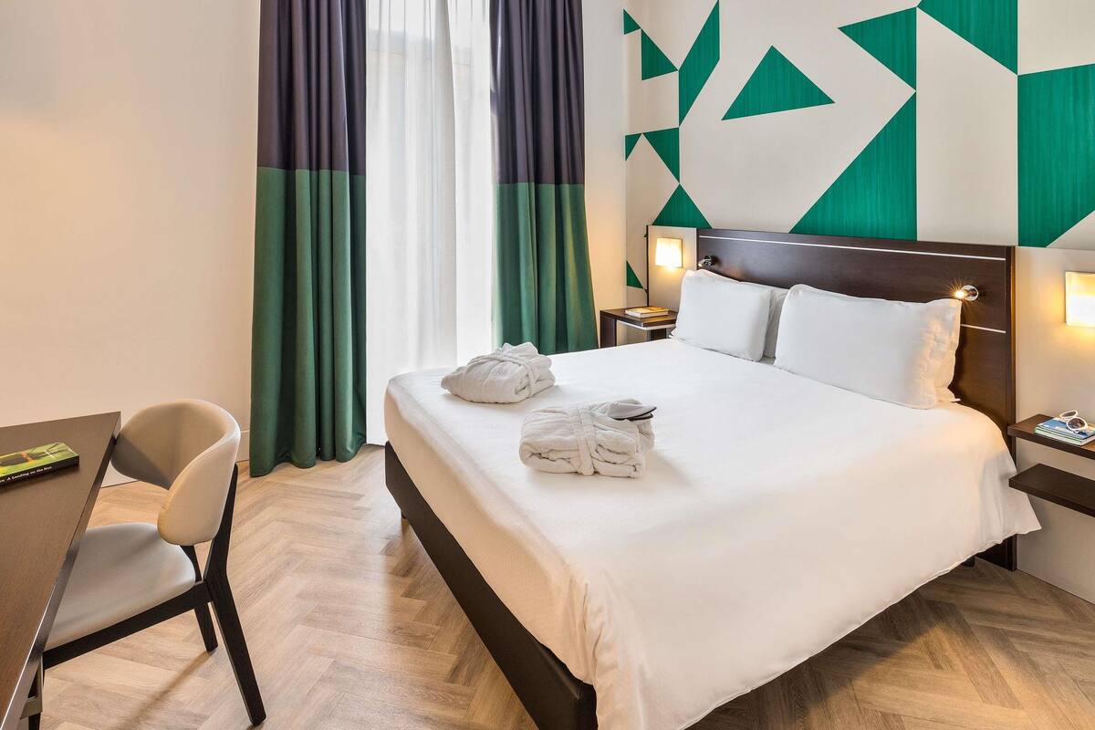 Quarto de hotel com uma cama bem arrumada, uma cadeira, mesa de cabeceira com abajur e parede com padrão geométrico.