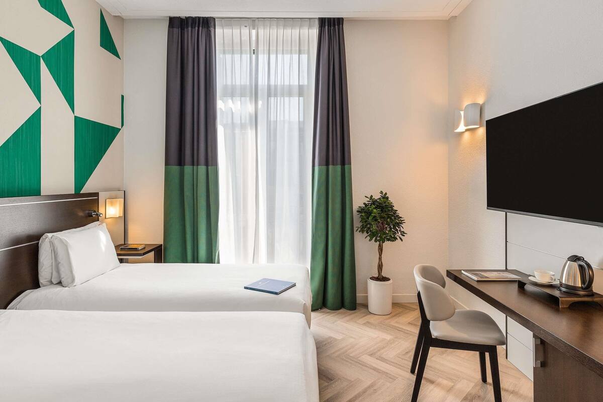 Quarto de hotel com cama, escrivaninha e cadeira, televisão, planta em vaso e cortinas.