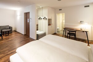 Quarto de hotel com uma cama grande, uma escrivaninha com cadeira, uma mesinha, um sofá e um guarda-roupa.