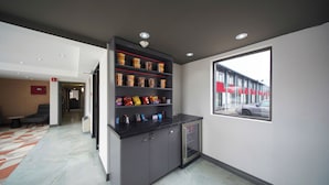 Um hall de hotel moderno com piso de mármore, um balcão de recepção escuro com distribuidores de lanches e vista para um prédio vermelho lá fora.
