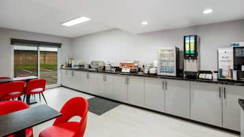 Cozinha moderna com geladeira, máquinas de café e micro-ondas. Há cadeiras vermelhas e uma mesa com tampo preto.
