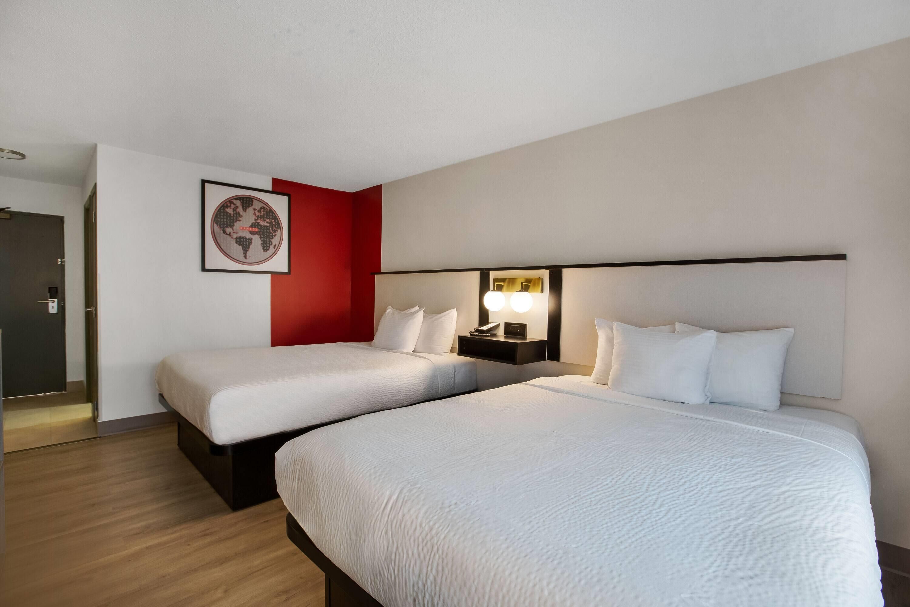 Quarto de hotel com duas camas, uma parede de destaque vermelha, um mapa-múndi emoldurado e uma luminária de cabeceira.