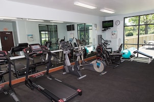 Uma academia com diversos equipamentos de exercícios, janelas amplas e uma saída de emergência visível.
