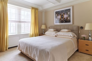 Uma cama grande com colcha branca, duas mesas de cabeceira com abajures, uma obra de arte emoldurada sobre a cama e uma janela com cortinas amarelas.