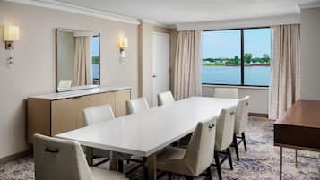 Uma sala de conferências de hotel com uma mesa grande, seis cadeiras, um minibar, um espelho e vista para um corpo de água.