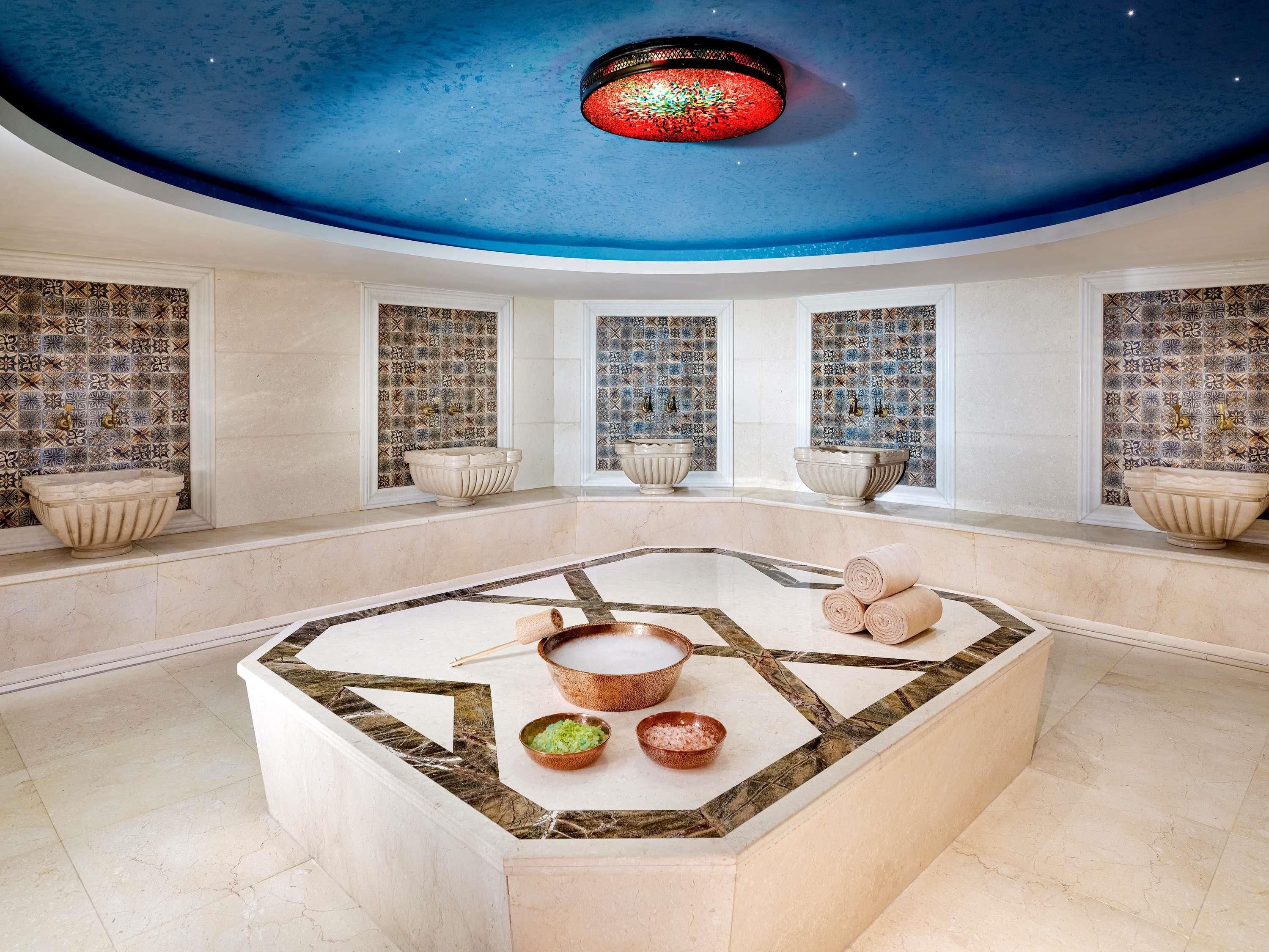 Um quarto de spa com uma banheira de mármore central, taças decorativas e paredes com azulejos estampados.