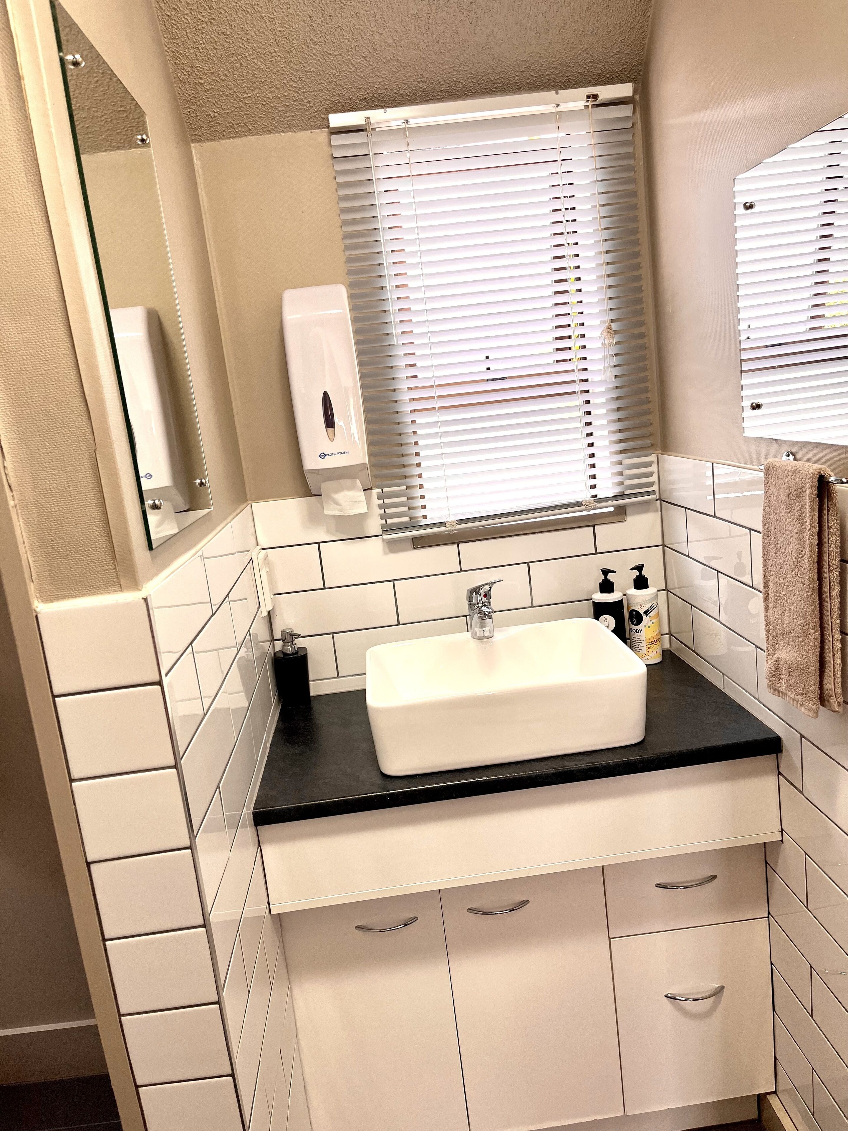 Um banheiro com pia branca, bancada preta e azulejos bege. Há um espelho, porta-toalhas e uma janela com persianas.