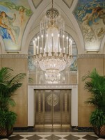 Um hall imponente com um lustre ornamentado, um grande mural e um elevador clássico.