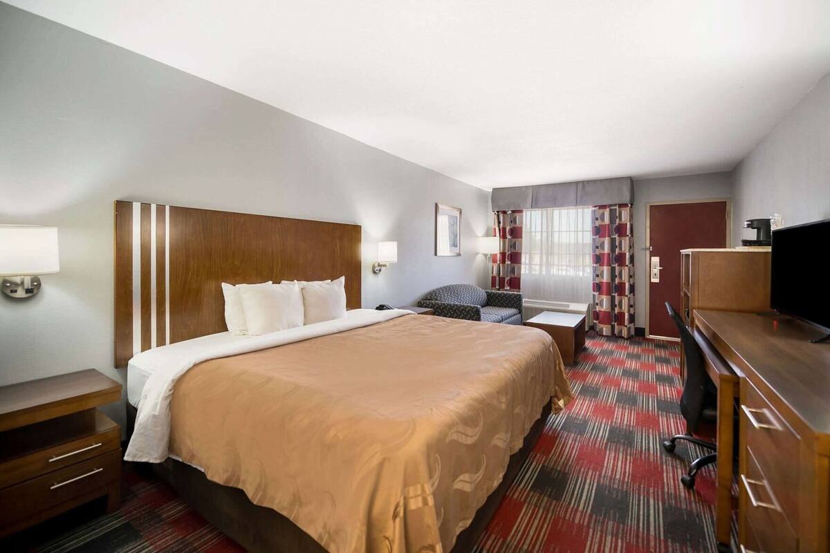 Quarto de hotel com uma cama grande, escrivaninha, cadeira, televisão e janela com cortinas.