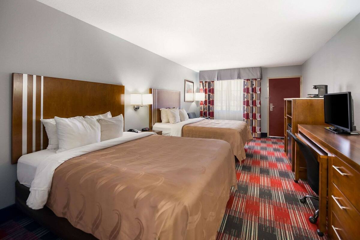 Quarto de hotel com duas camas, uma escrivaninha, uma televisão e um tapete estampado.