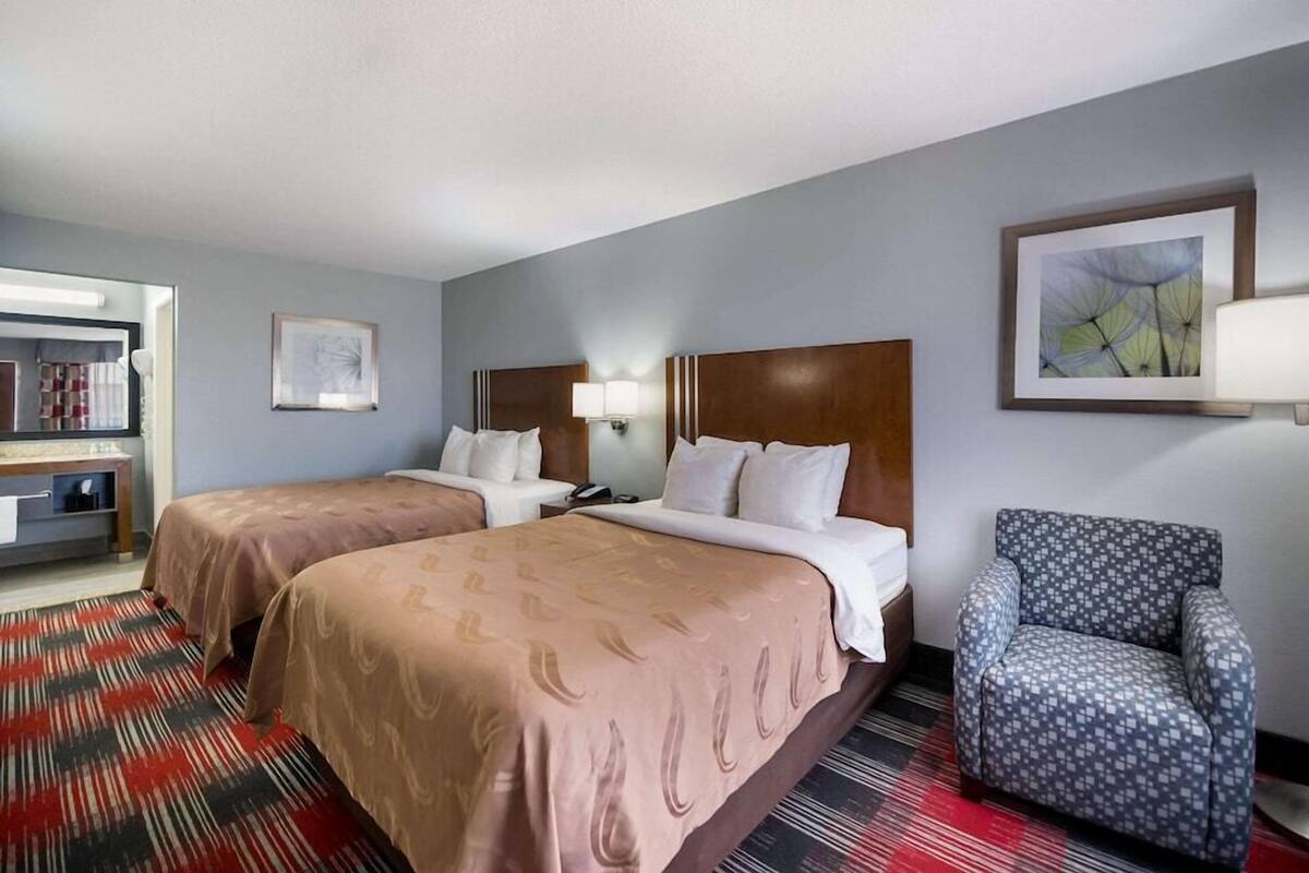 Quarto de hotel com duas camas, uma cadeira, uma escrivaninha e um espelho.