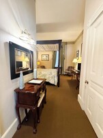 Quarto de hotel com cama, escrivaninha, cadeira, luminária, espelho e porta.