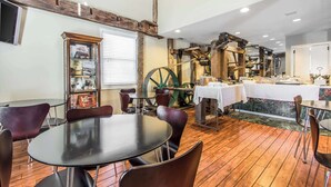 Restaurante com piso de madeira, mesas redondas e cadeiras. Há uma grande roda e um balcão com toalha de mesa branca.
