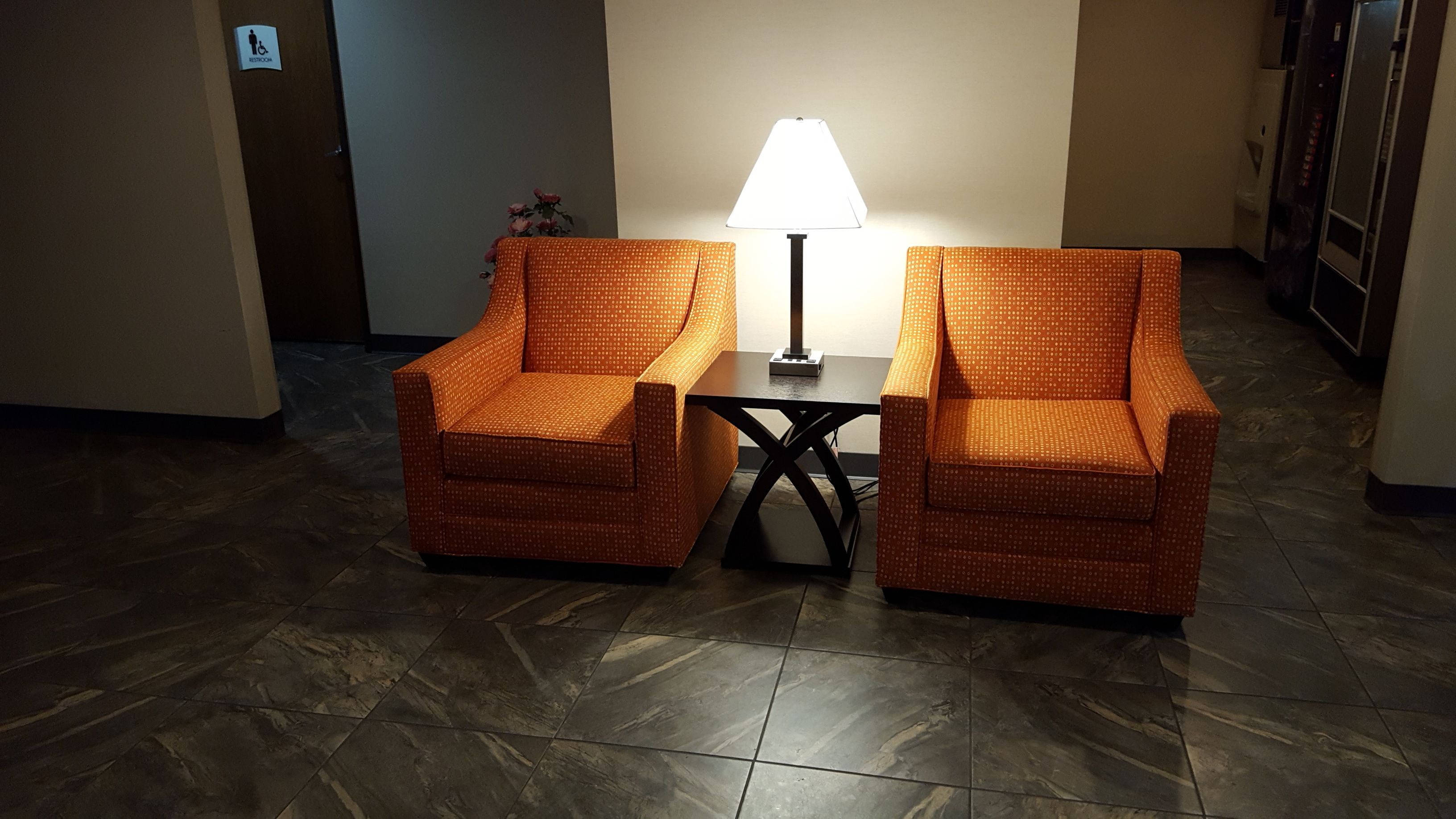 Um saguão de hotel com dois poltronas laranja, uma mesa com abajur, um sinal de banheiro e piso ladrilhado.