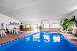 Uma piscina interior com água azul cristalina, rodeada por espreguiçadeiras e mesas brancas.