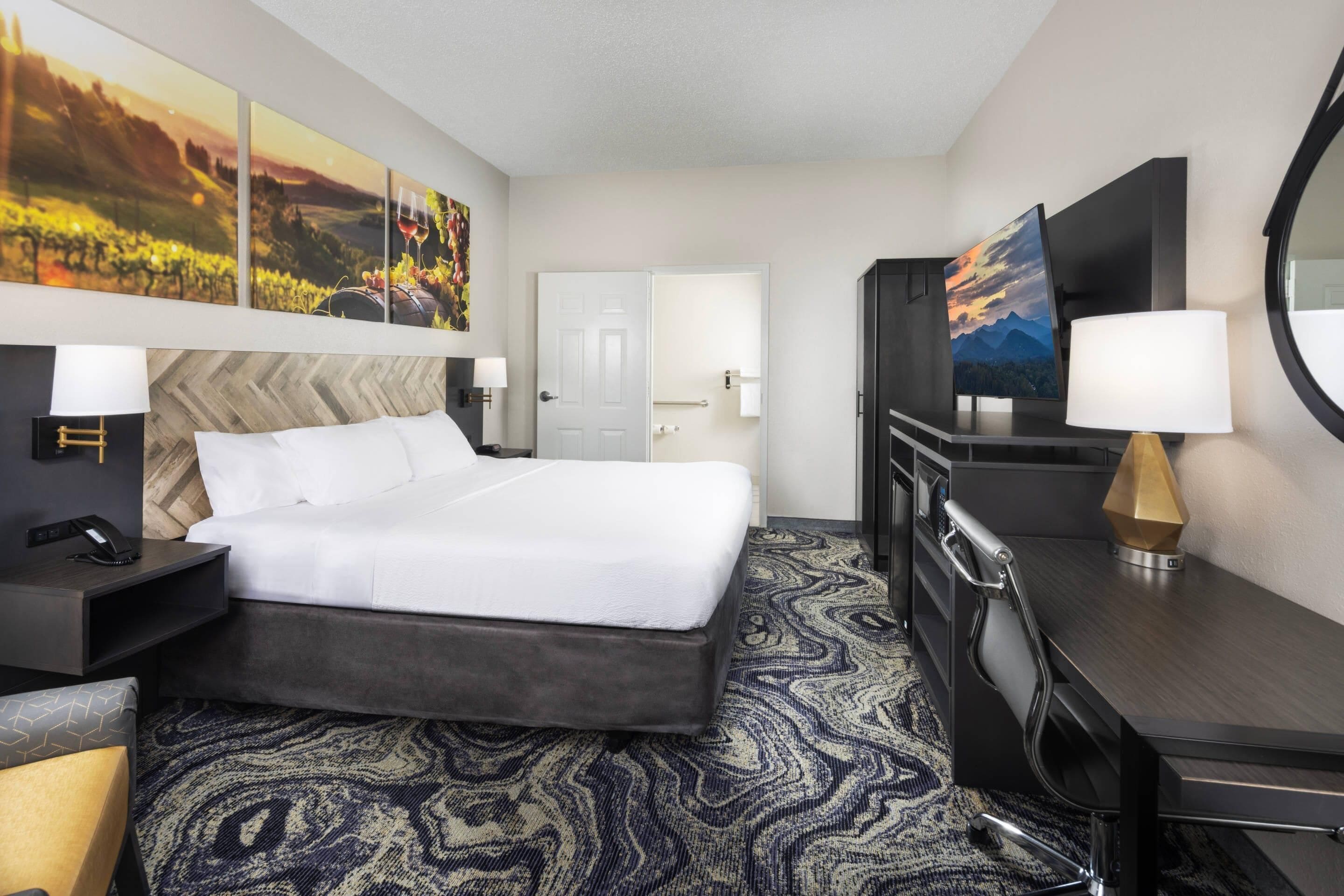 Quarto de hotel com uma cama grande, uma escrivaninha com cadeira, cómoda, espelho e obras de arte nas paredes.