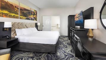 Quarto de hotel com uma cama grande, uma escrivaninha com cadeira, cómoda, espelho e obras de arte nas paredes.