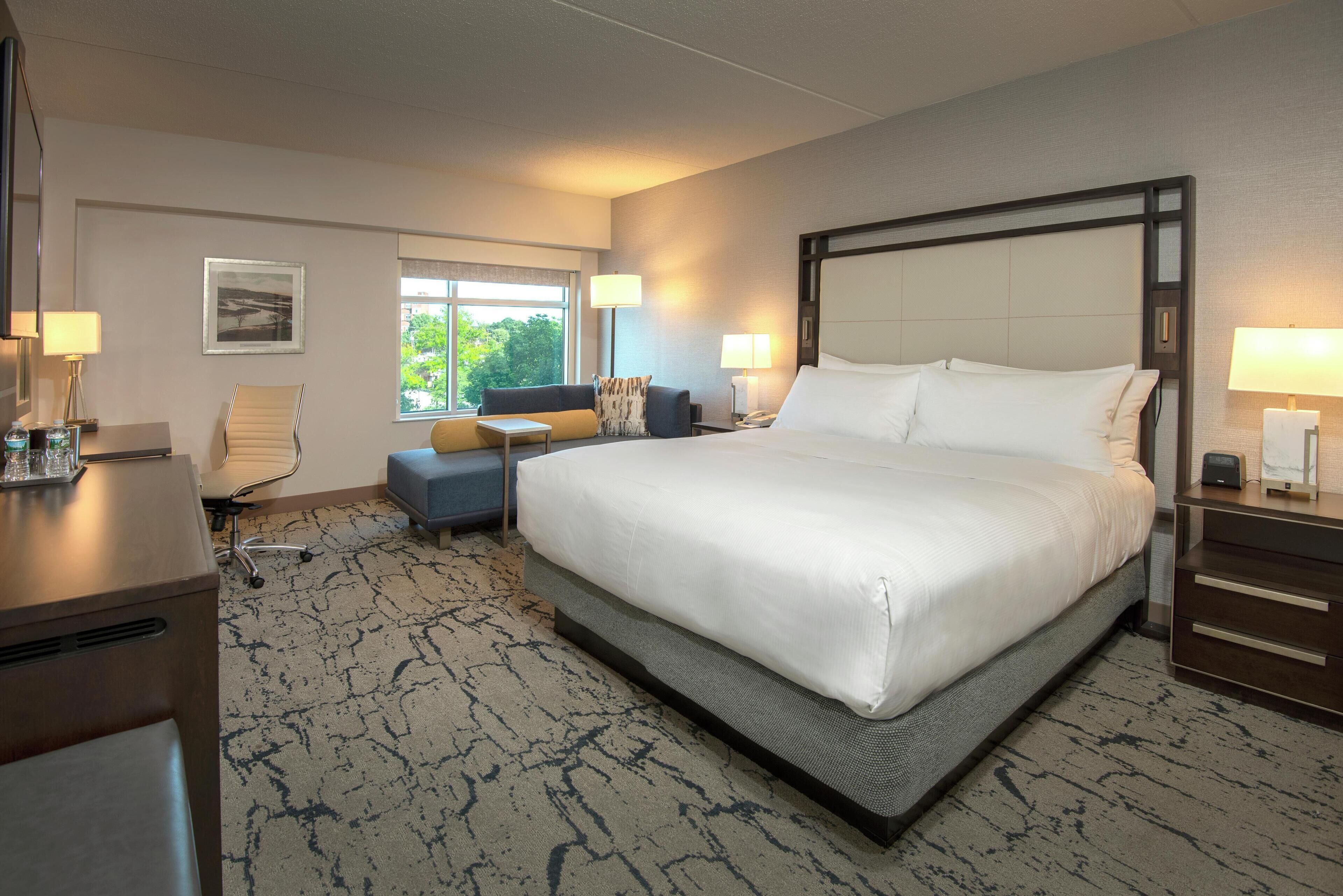 Quarto de hotel com uma cama grande, uma escrivaninha, uma cadeira, um sofá e uma janela.
