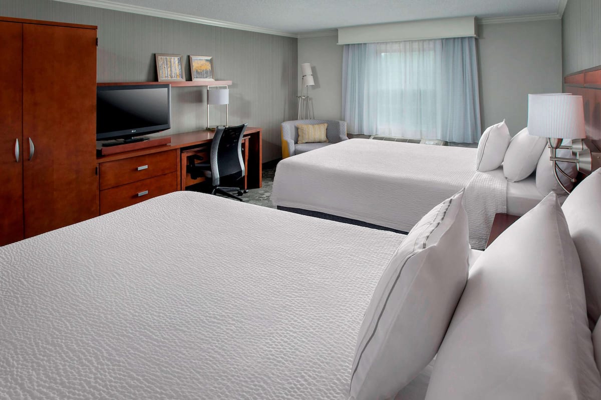 Quarto de hotel com duas camas, uma escrivaninha com televisão, uma cadeira e um abajur.