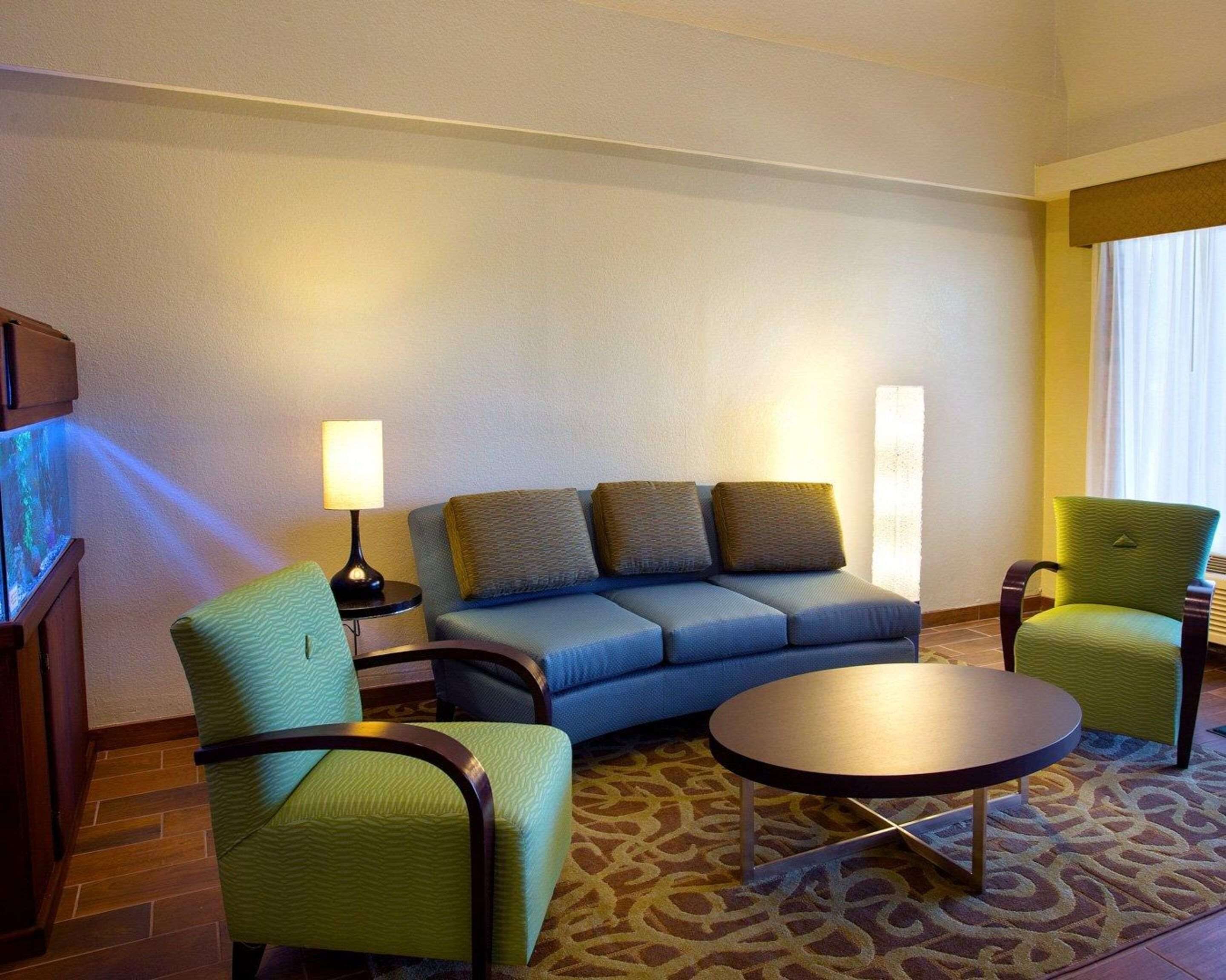 Quarto de hotel com sofá azul, poltrona verde, mesa de centro redonda e uma luminária.