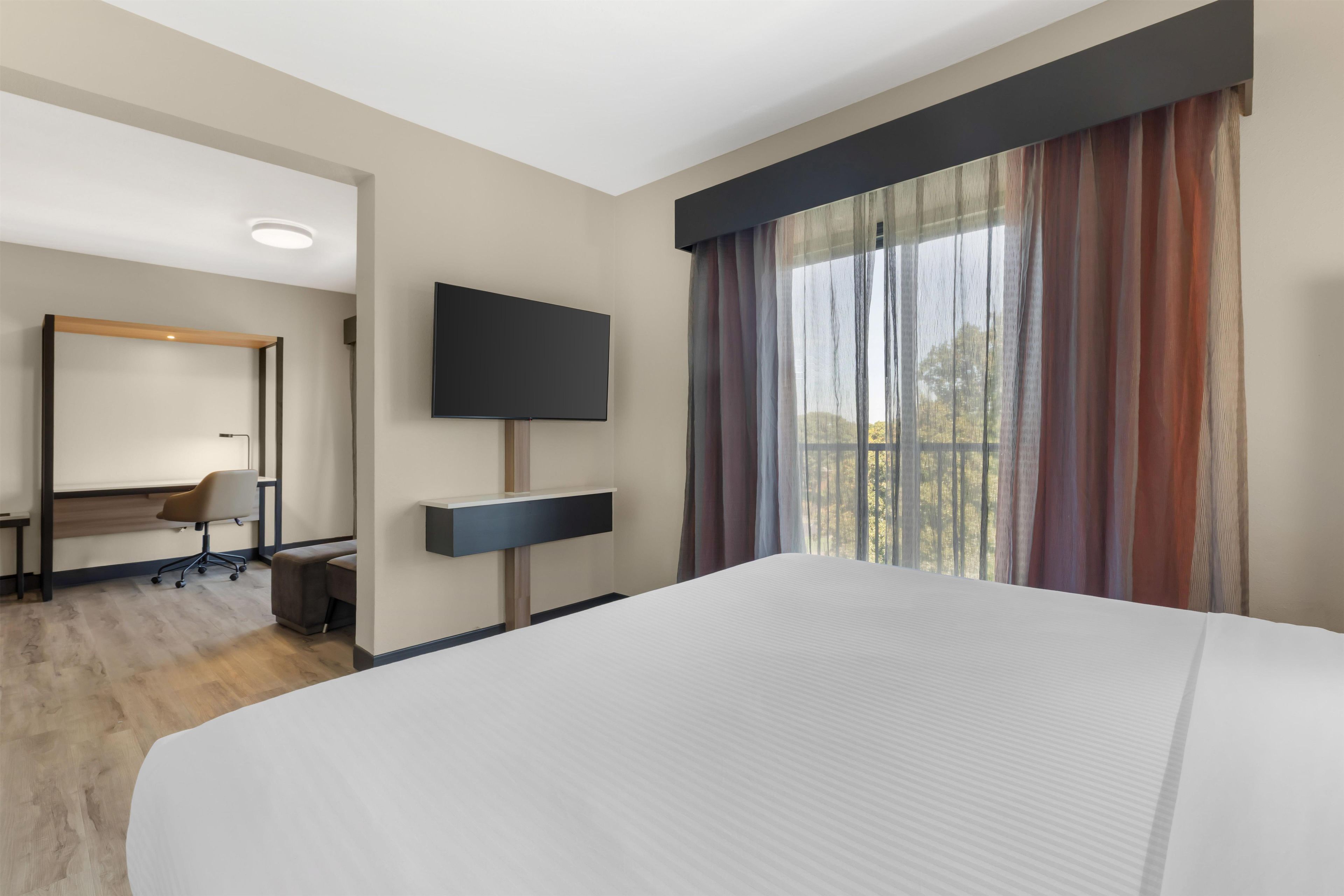 Quarto de hotel com uma cama grande, uma escrivaninha com cadeira, televisão de tela plana na parede e uma janela com cortinas.