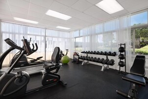 Uma academia com equipamentos de cardio, estações de musculação e uma grande janela com luz natural.