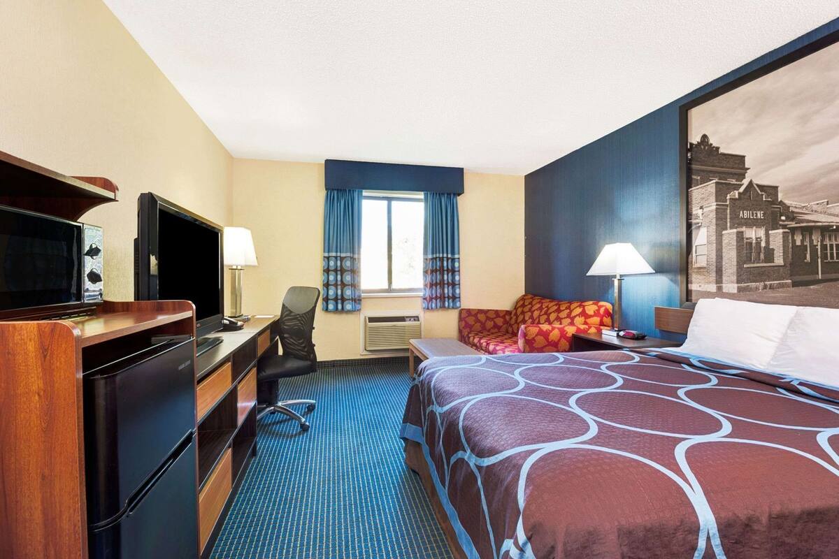 Quarto de hotel com cama, escrivaninha com televisão, cadeira, janela com cortinas e um mural na parede.