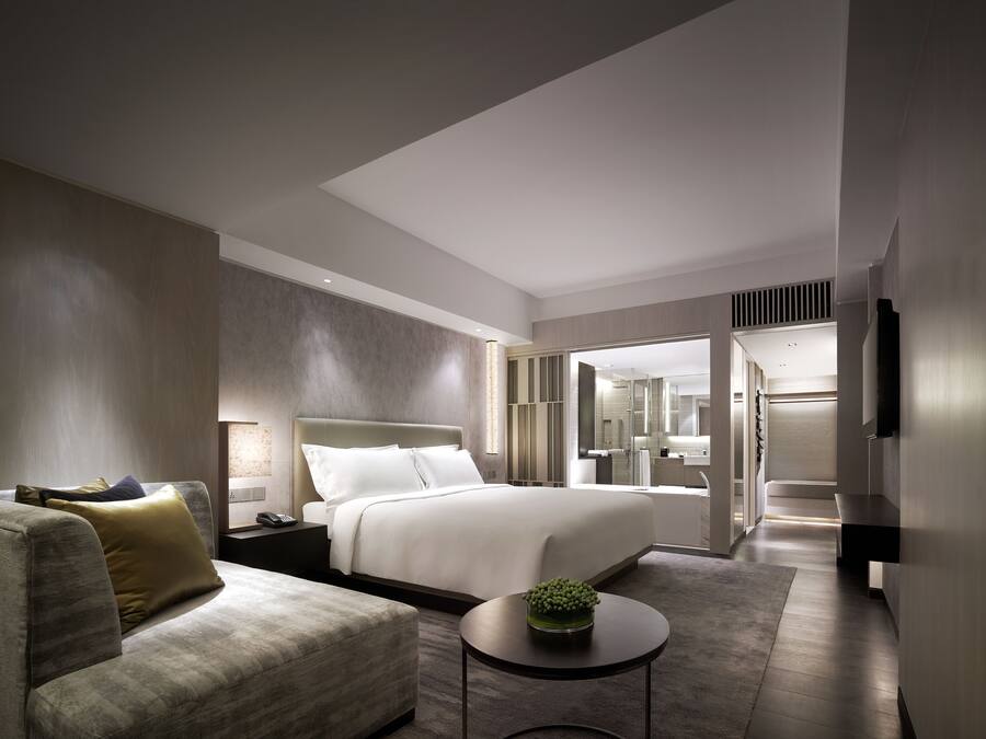 Quarto de hotel moderno com uma cama grande, um sofá, uma mesa redonda pequena com uma planta e um banheiro visível por uma porta aberta.