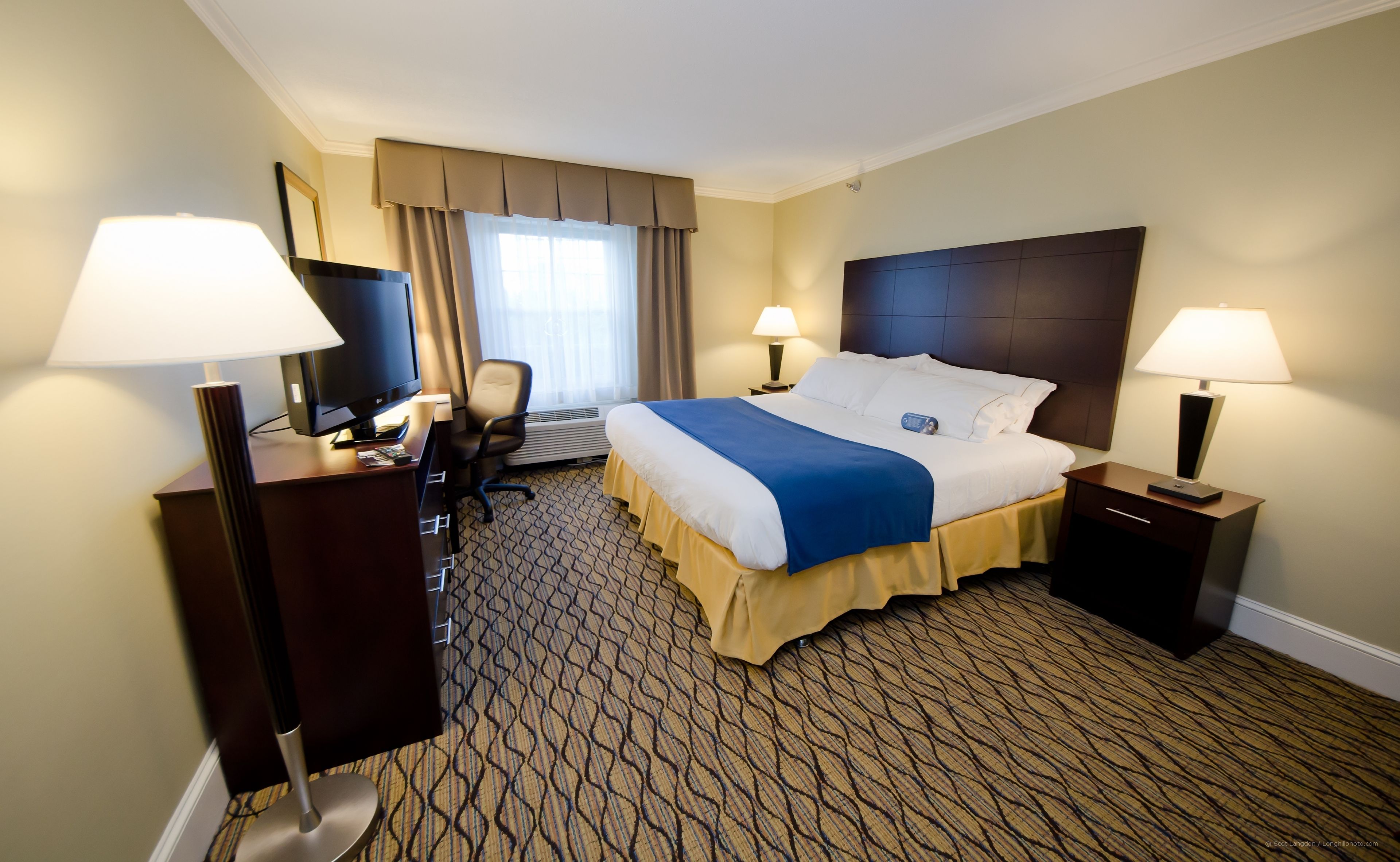 Quarto de hotel com cama king-size, duas mesas de cabeceira com abajures, escrivaninha com televisão e computador, uma cadeira e carpete estampado.
