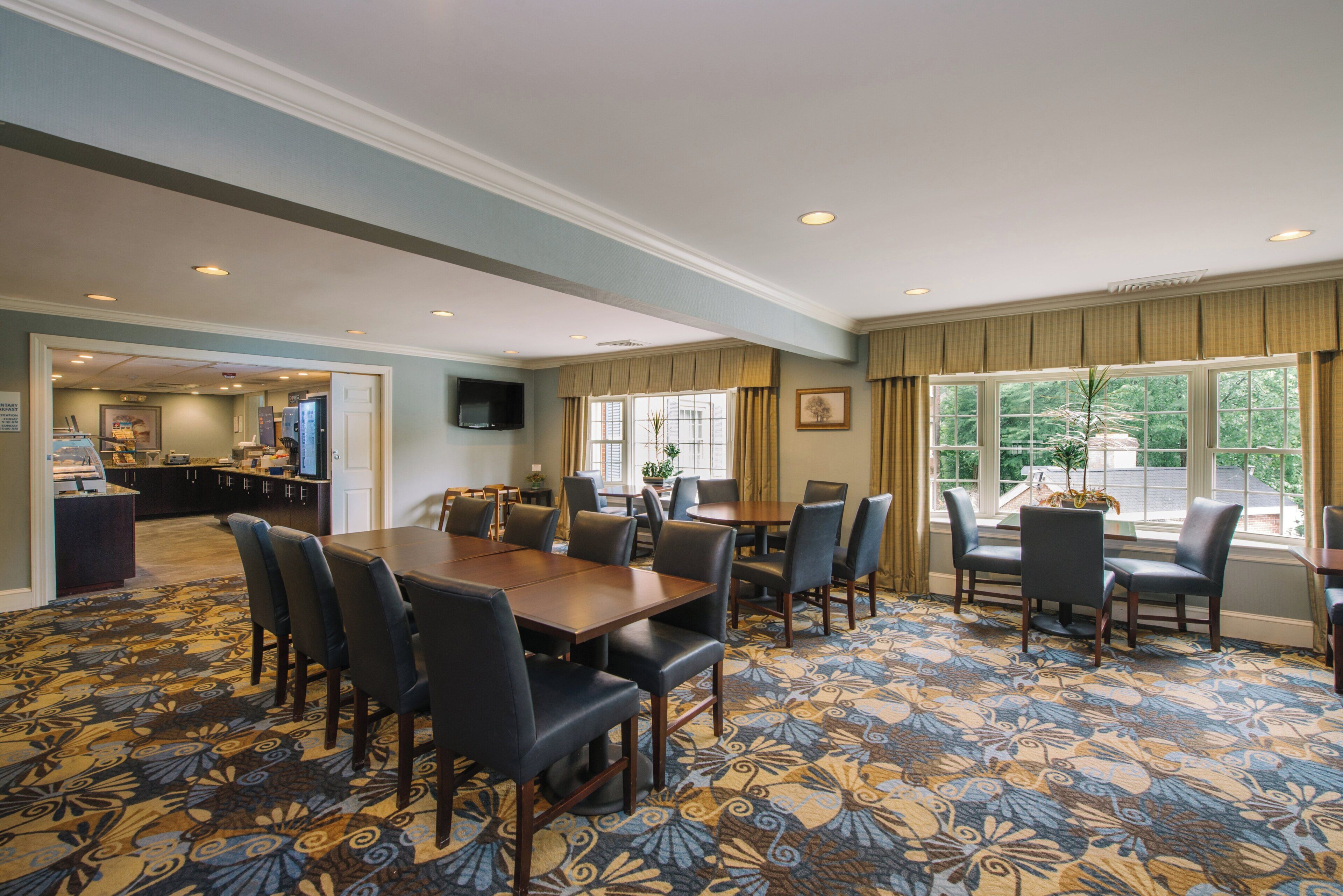 Um espaço de refeições de hotel com uma mesa grande e cadeiras, uma cozinha ao fundo e vista para o exterior com vegetação.
