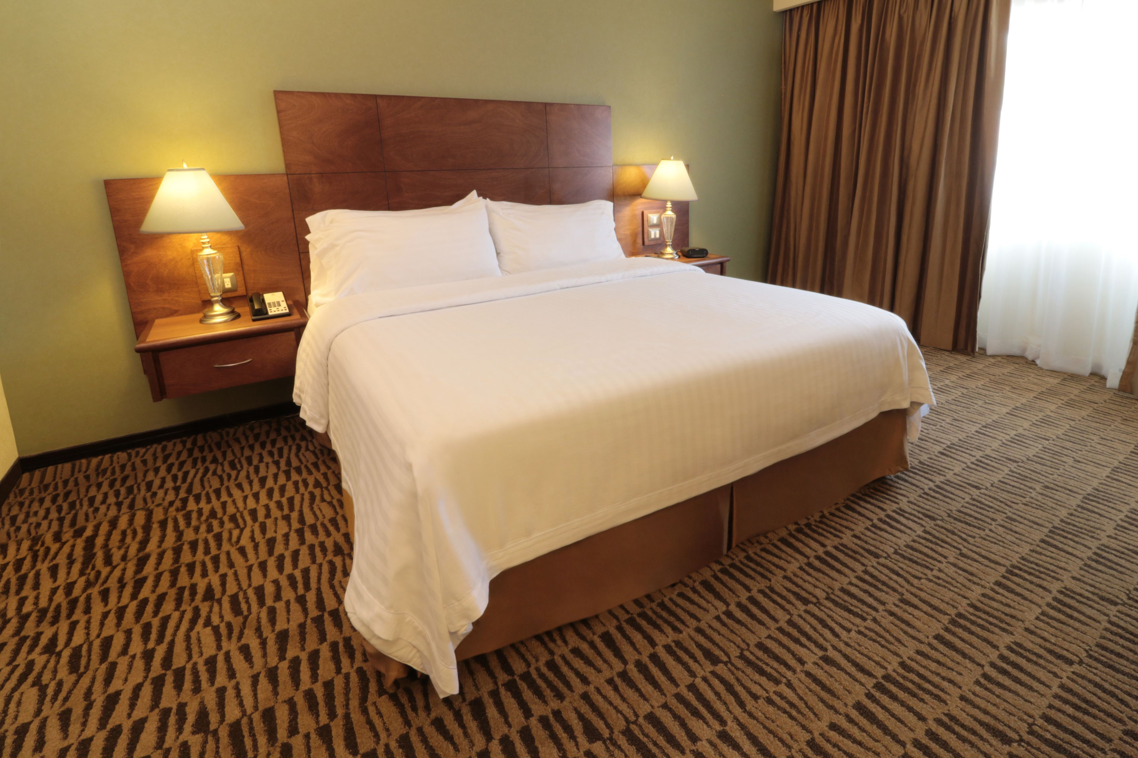 Quarto de hotel com uma cama grande, duas mesas de cabeceira com luminárias, uma cabeceira de madeira e um tapete estampado.