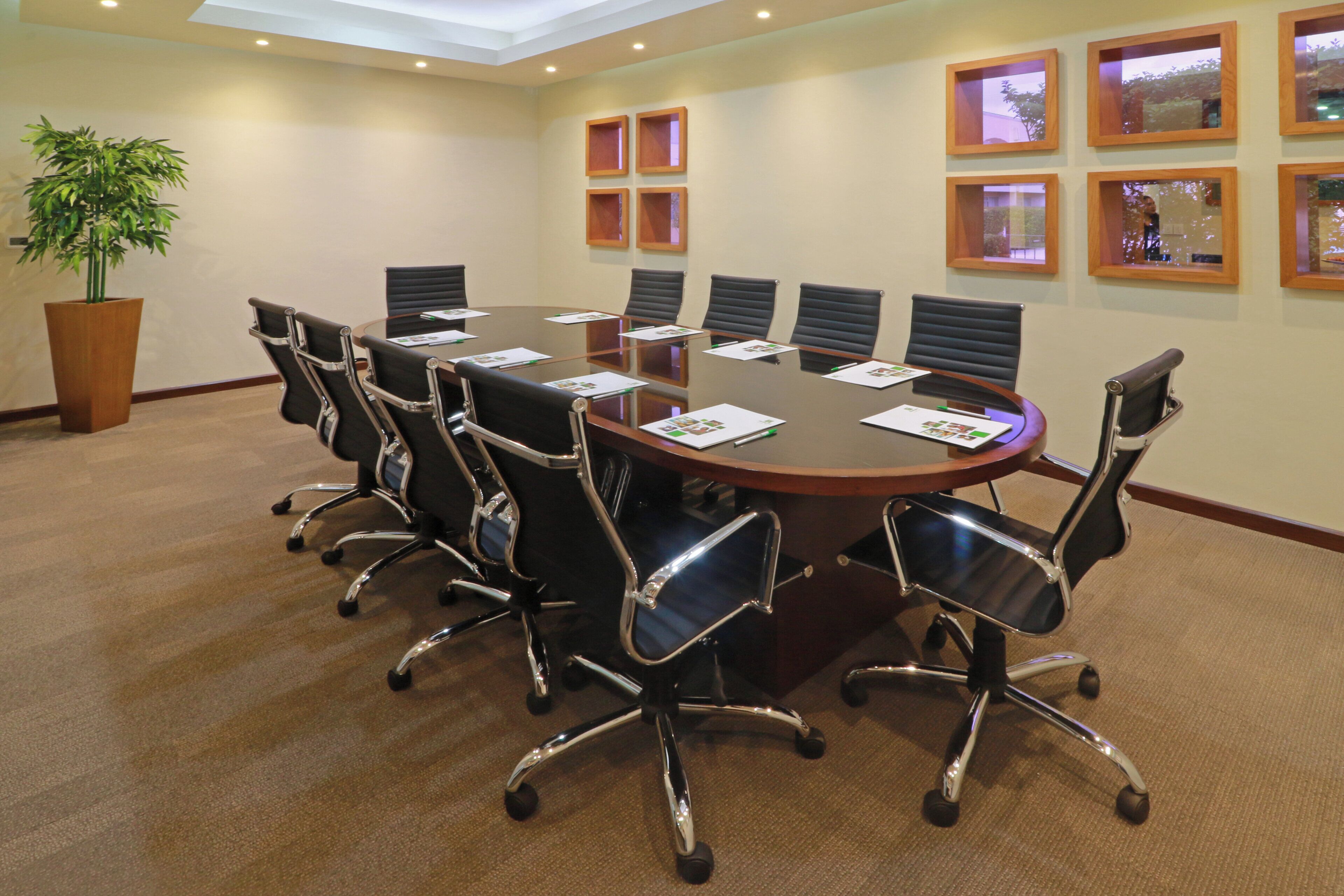 Sala de conferência com mesa redonda, seis cadeiras e um vaso de plantas.