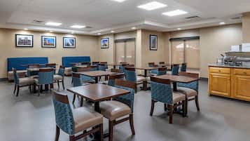 Um espaço de refeições de hotel com várias mesas e cadeiras, uma área de cozinha e quadros nas paredes.