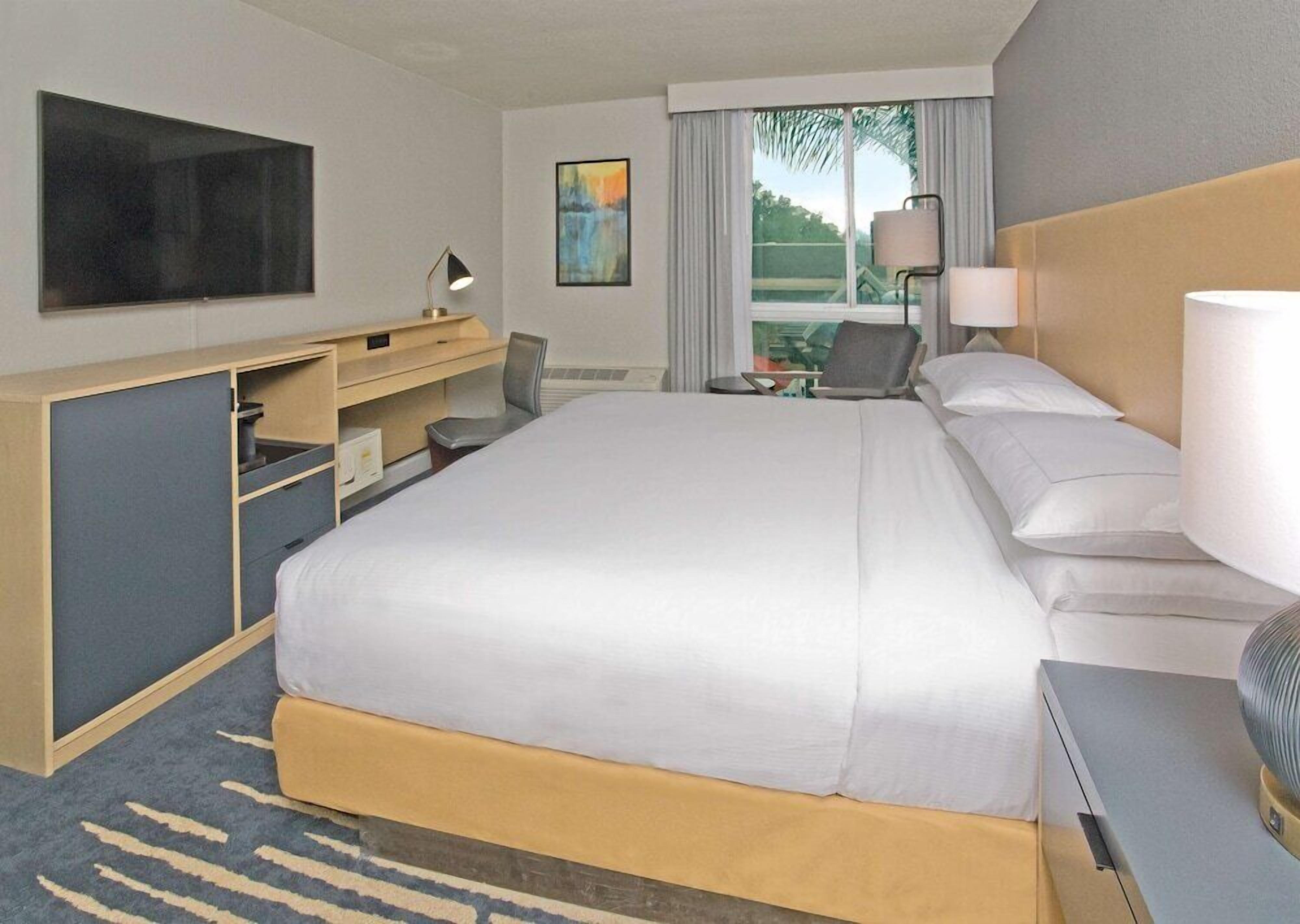 Quarto de hotel com uma cama grande, uma mesa com cadeira, televisão, pintura e janela com cortinas.