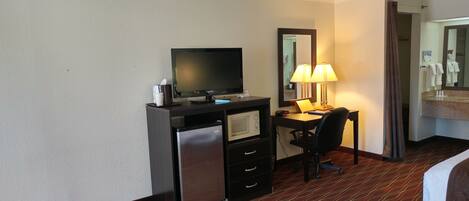 Quarto de hotel com escrivaninha, cadeira, televisão, micro-ondas e um pequeno frigorífico.