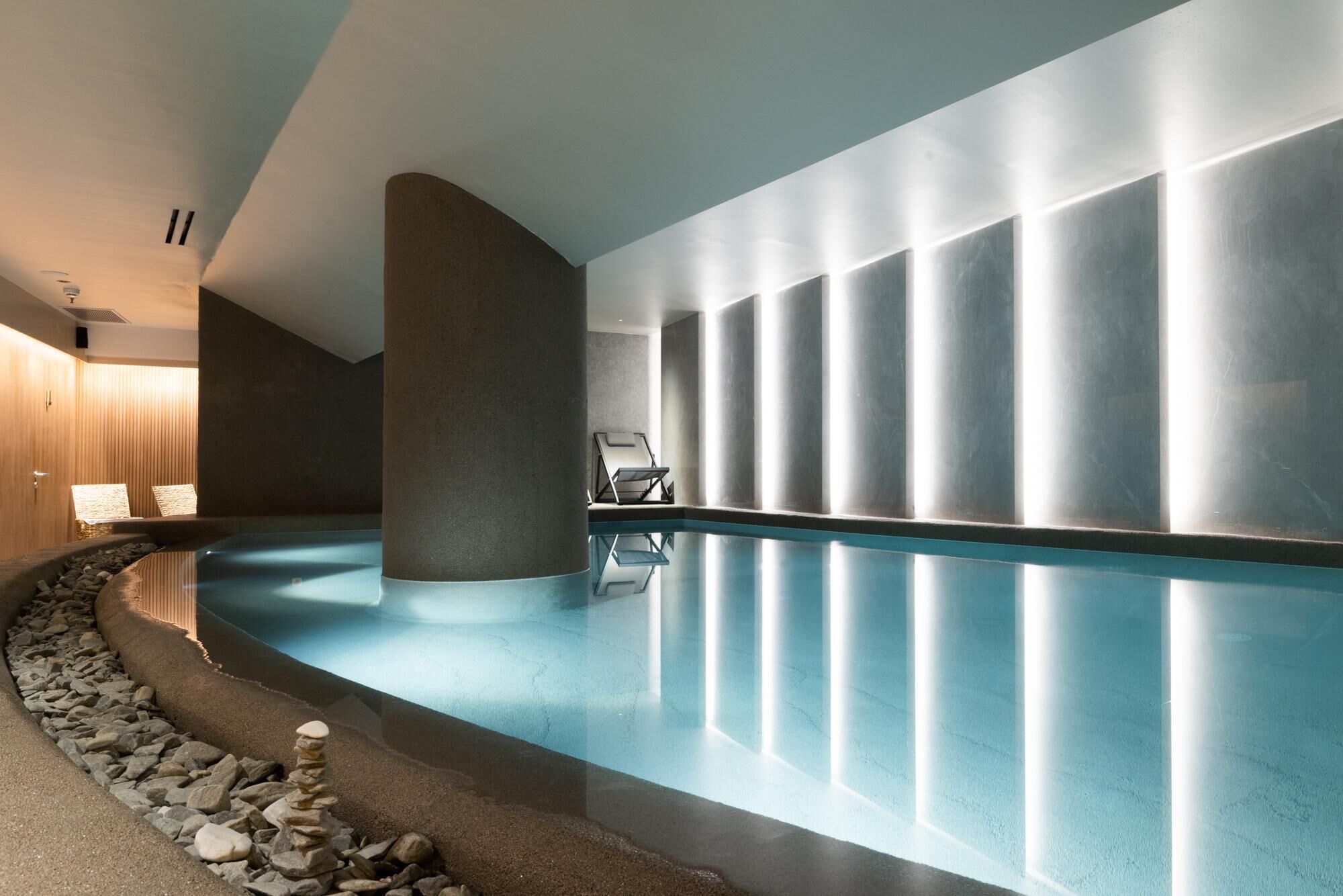 Piscina interior moderna com superfície aquática límpida, borda revestida em pedra e design minimalista, com um pilar imponente e fileira de luzes ao longo da parede.